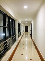 Um corredor longo e bem iluminado, com chão de azulejos, grades de metal e uma sequência de portas no final.