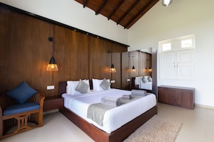 Quarto com uma cama grande, cabeceira de madeira, duas luminárias pendentes, um banco de madeira, um espelho grande e um guarda-roupa branco.