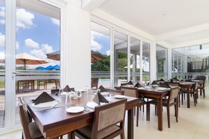 Uma área de refeições com mesas preparadas para uma refeição, grandes janelas e vista para o exterior.