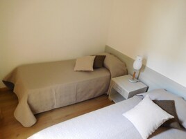 Quarto pequeno e simples com duas camas de solteiro, uma mesa de cabeceira com abajur e piso de madeira.
