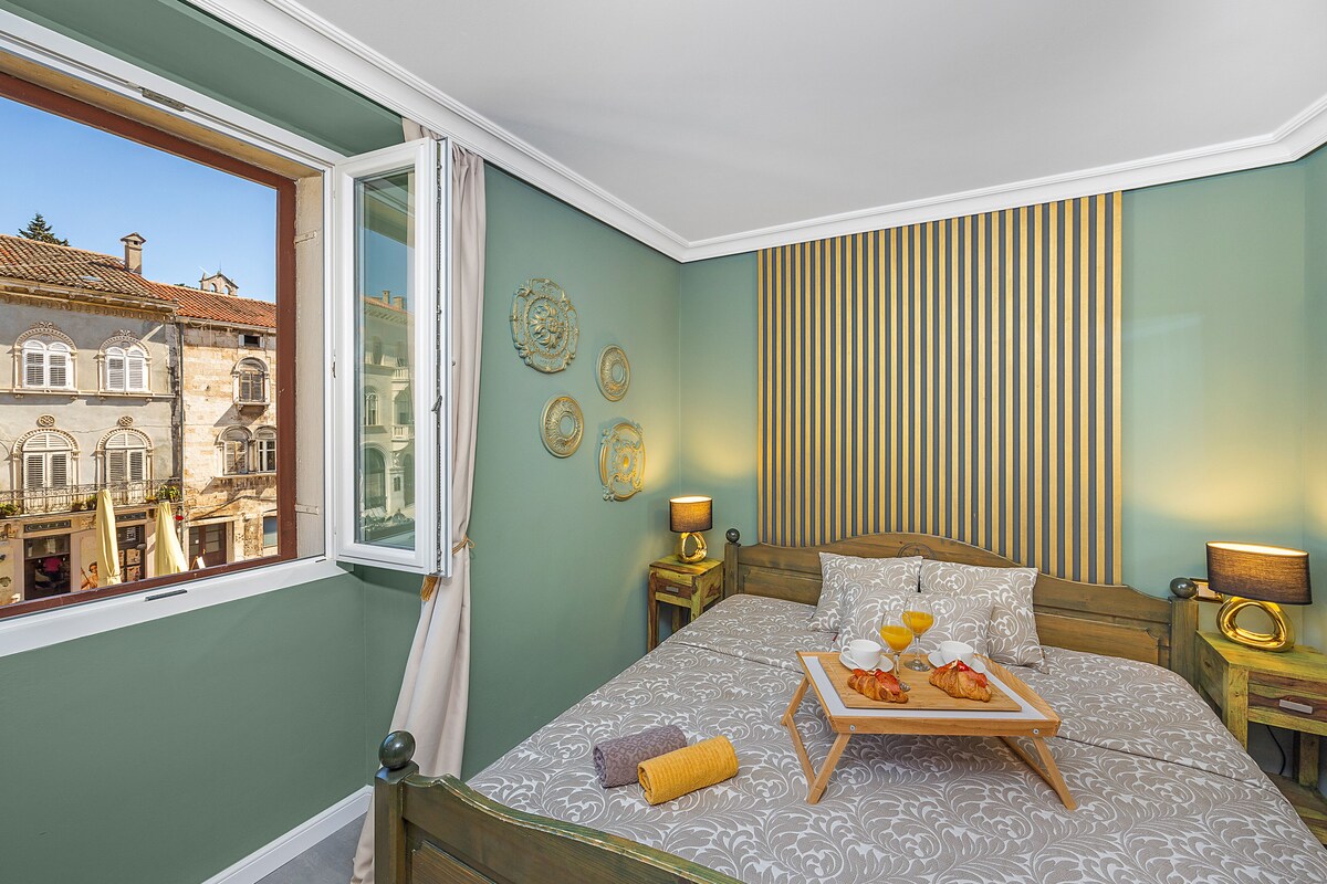 Quarto com cama, mesa de cabeceira, janela e vista para edifícios.