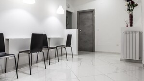 Uma área de espera com paredes brancas, uma porta cinzenta e um radiador. Há quatro cadeiras pretas e duas luminárias pendentes.