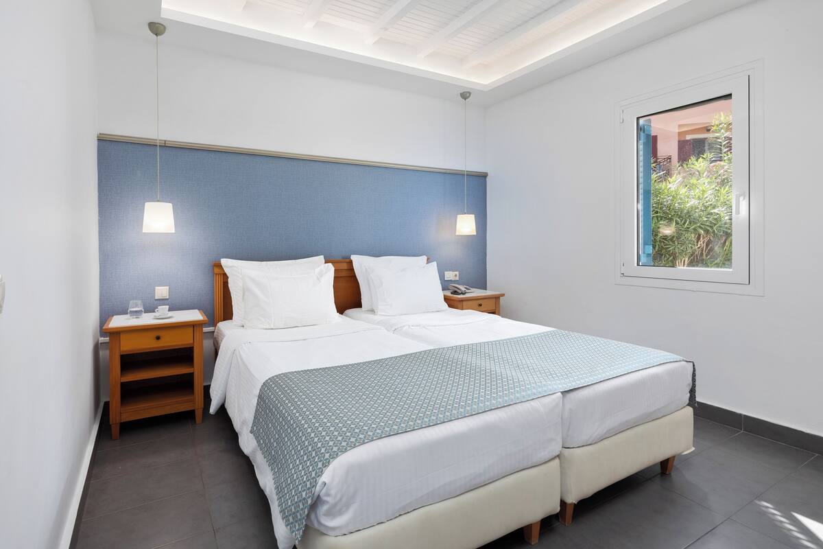 Uma cama de casal com colcha estampada em azul e branco, duas mesas de cabeceira com abajures e uma janela com vista para o verde.