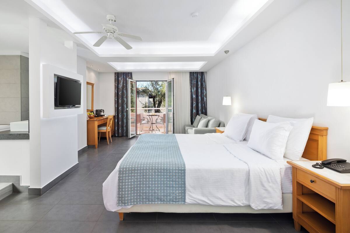 Quarto de hotel com cama grande, escrivaninha, televisão e varanda com cortinas.