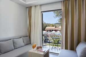 Quarto de hotel com sofá, uma pequena mesa com um livro e copos de suco de laranja, e vista para uma piscina e área de estar ao ar livre.