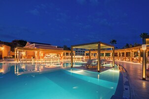 Piscina com espreguiçadeiras e área de bar, cercada por edifícios com iluminação acolhedora.