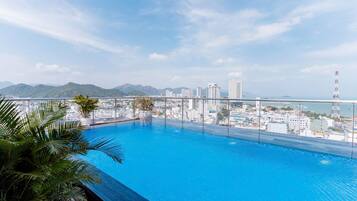 Piscina infinita no terraço com vista para a cidade, palmeiras e céu claro.