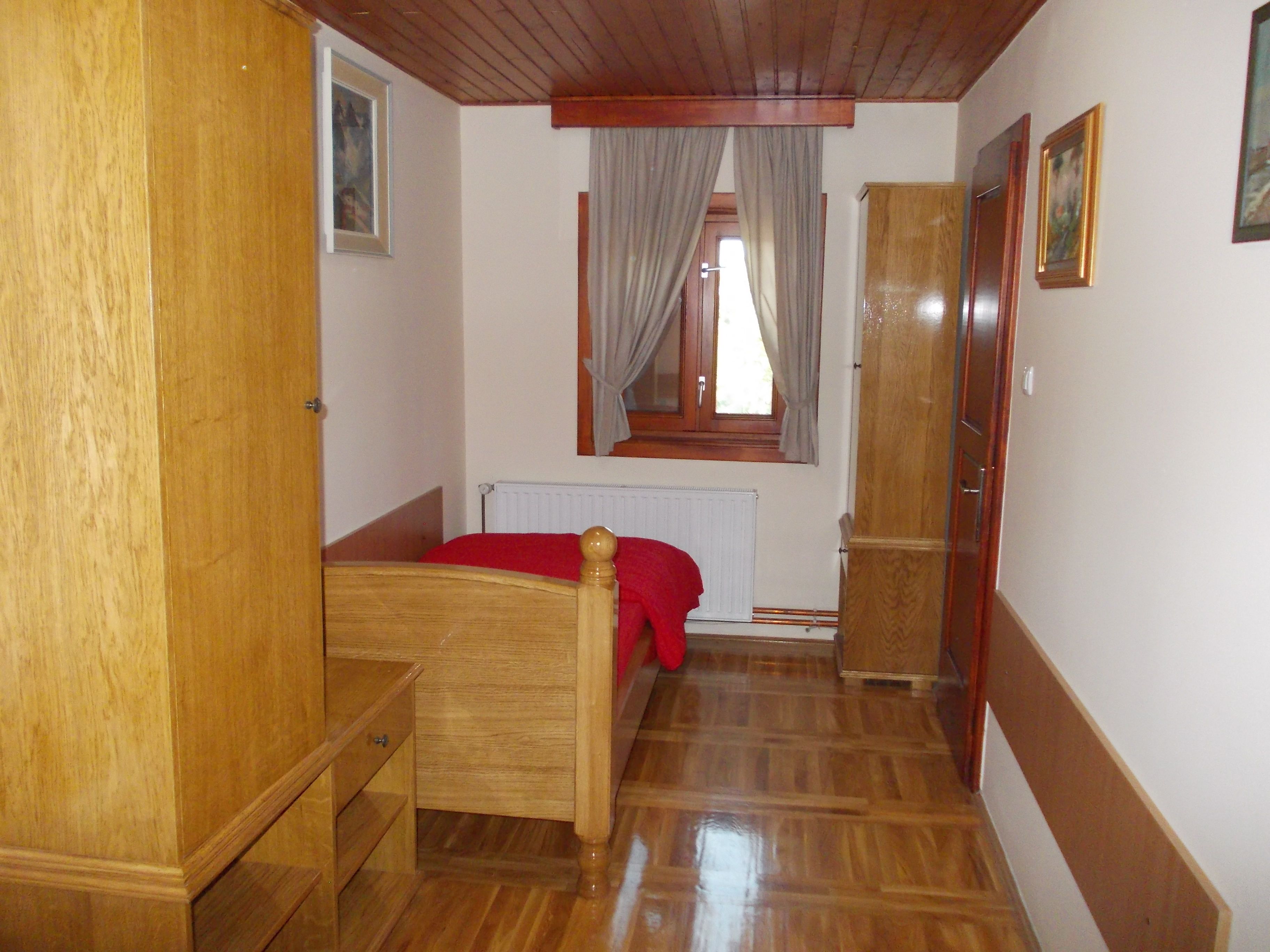 Quarto com estrutura de madeira, uma cama, guarda-roupa de madeira, radiador e janela com cortinas.