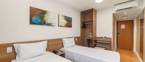 Quarto de hotel com duas camas de solteiro, cabeceira de madeira, escrivaninha com cadeira, guarda-roupa e arte abstrata na parede.