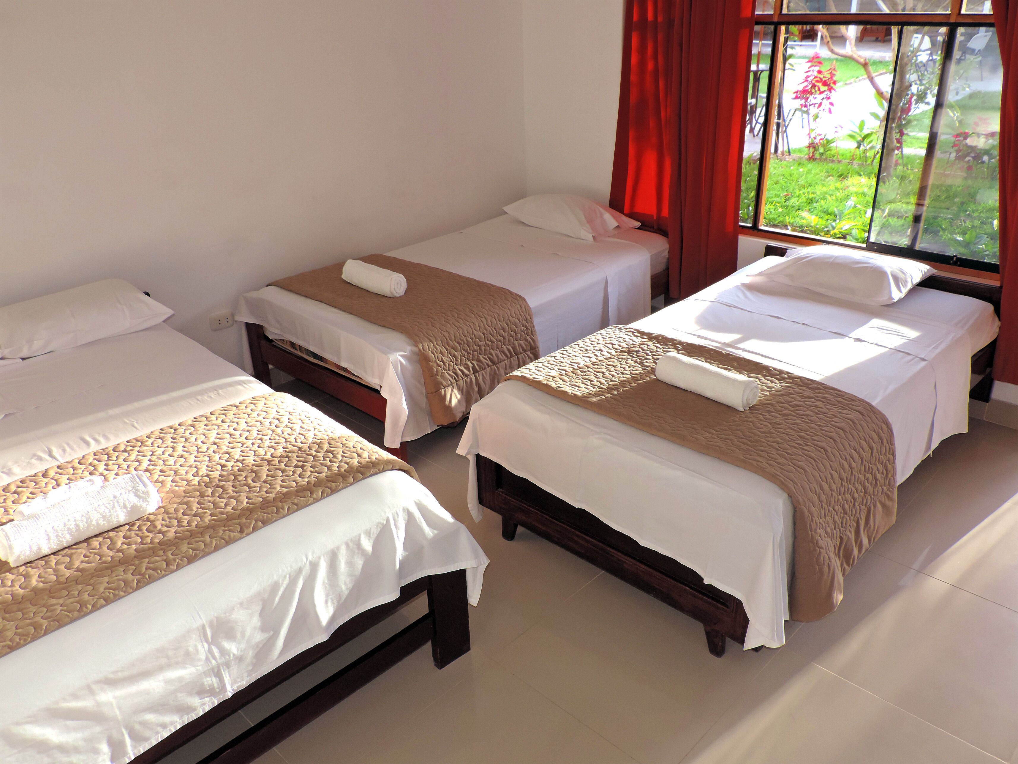 Quarto de hotel com três camas de solteiro, cada uma com edredom branco e bege e uma toalha enrolada.