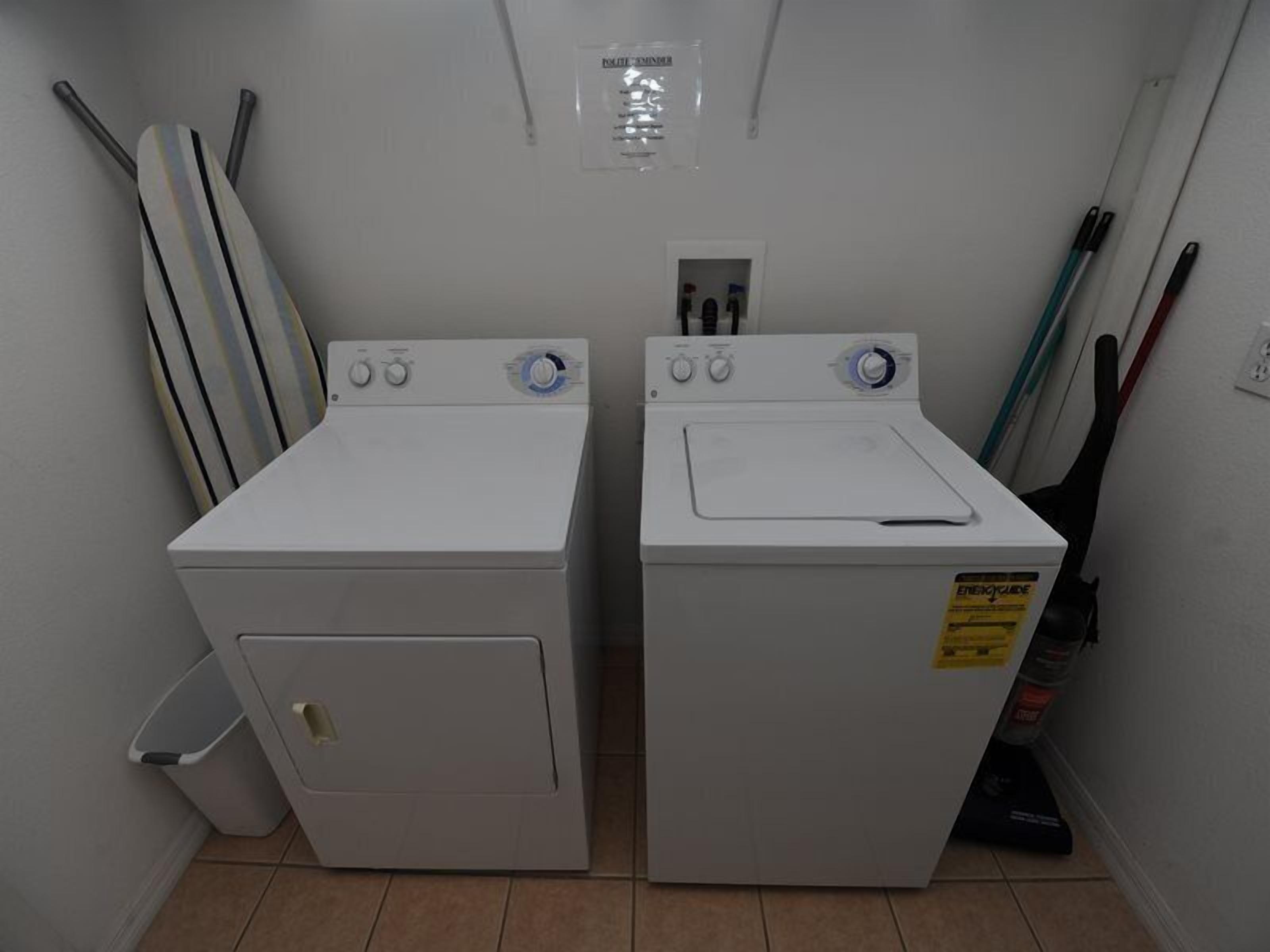 Uma lavanderia com duas máquinas de lavar brancas, uma lixeira e uma tomada.