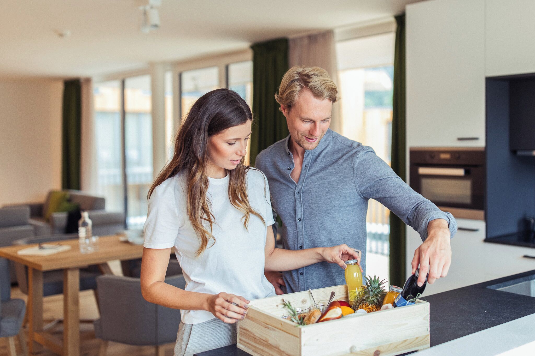 Cozinha moderna com uma caixa de madeira para frutas e legumes no balcão, um homem e uma mulher ao lado dela, e uma área de jantar ao fundo.