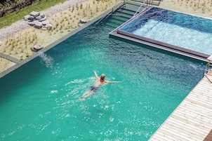 Uma pessoa nada em uma piscina azul cristalina, com um deck de madeira e uma pequena área de jardim com pedras e plantas.