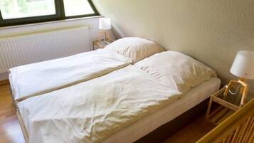 Uma cama de casal com roupa de cama branca em um quarto com uma janela grande, um radiador e uma mesinha de cabeceira com abajur.