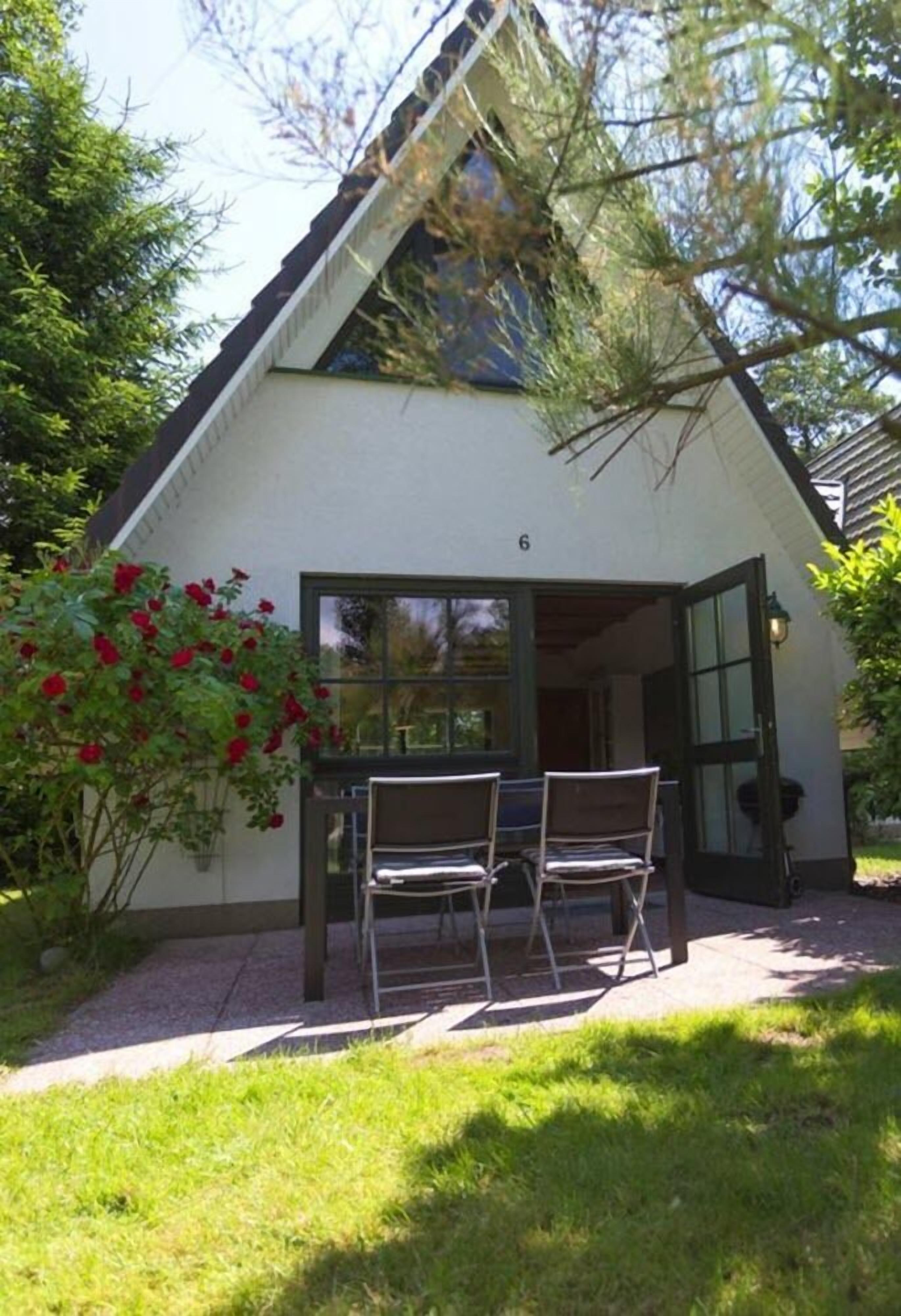 Uma casa pequena, térrea, com telhado inclinado, o número '6' na fachada e um alpendre com duas cadeiras.