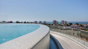 Uma piscina com borda curva, cercada por guarda-vidro, com vista para um horizonte costeiro com edifícios e o mar.