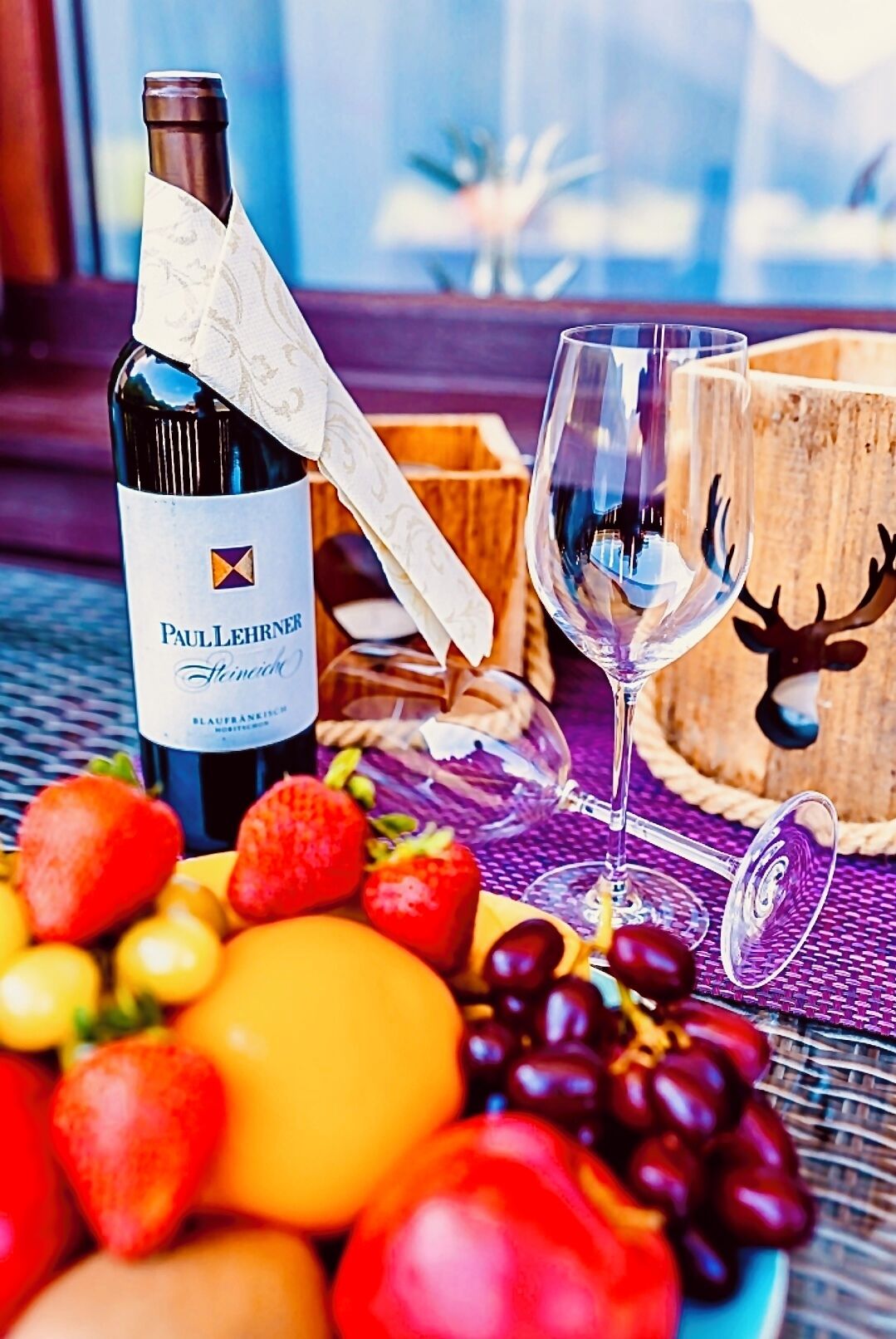 Uma garrafa de vinho, uma taça e um prato de frutas sobre uma mesa com um jogo americano de palha.