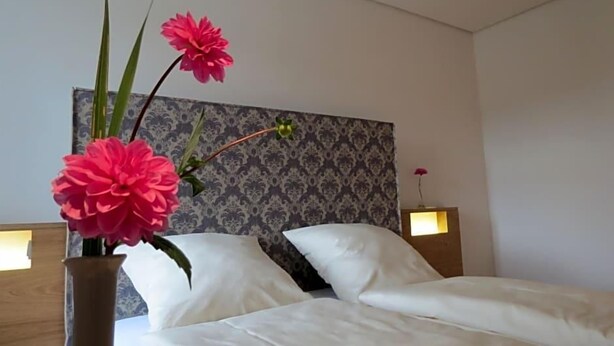 Quarto de hotel com cama, roupa de cama branca, um vaso com flores e um painel de cabeceira com papel de parede estampado.