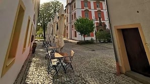 Pátio de calçamento com um edifício com duas torres verdes em formato de cúpula, uma pequena árvore e mesas ao ar livre.