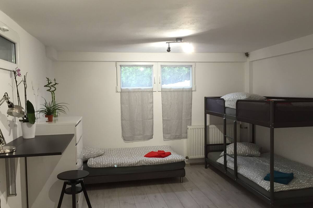Quarto compacto com uma cama beliche, uma pequena escrivaninha com luminária, um radiador e duas janelas com cortinas.