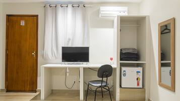 Quarto compacto com porta de madeira, uma escrivaninha branca com computador, cadeira e um pequeno frigorífico.