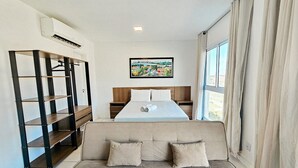 Quarto de hotel com cama, sofá, estante de livros, pintura e janela com cortinas.