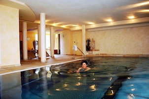 Uma piscina interior ampla com uma pessoa na água, rodeada por colunas e um chão em azulejo.