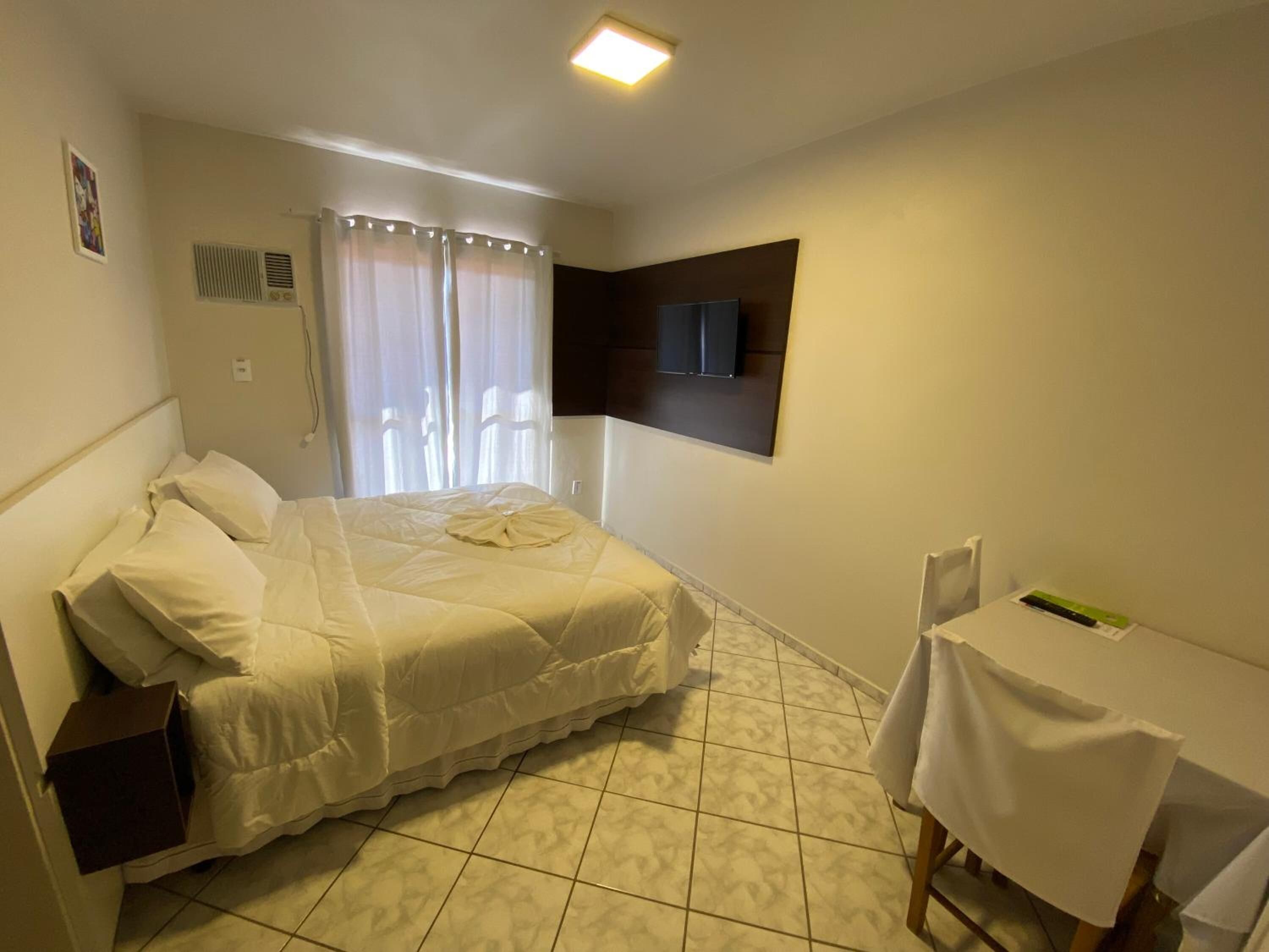 Quarto de hotel com cama, escrivaninha, cadeira, televisão e janela com cortinas.