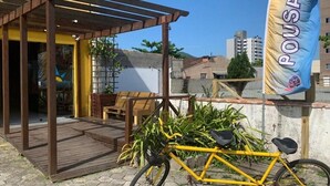Uma bicicleta amarela estacionada em frente a um edifício com pérgola de madeira e uma bandeira com a inscrição "POUSSA".