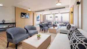 Um lobby de hotel moderno com sofás, poltronas e mesa de centro em tons de cinza. Há uma TV montada na parede, e o espaço possui grandes janelas com vista para edifícios.