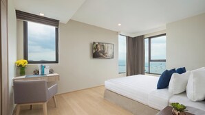 Quarto de hotel moderno com uma cama grande, uma escrivaninha com cadeira, uma mesinha de centro com uma planta e amplas janelas com vista para o mar.