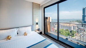 Quarto de hotel com uma grande janela, cama com almofadas, mesa de cabeceira com abajur e vista para a paisagem urbana.