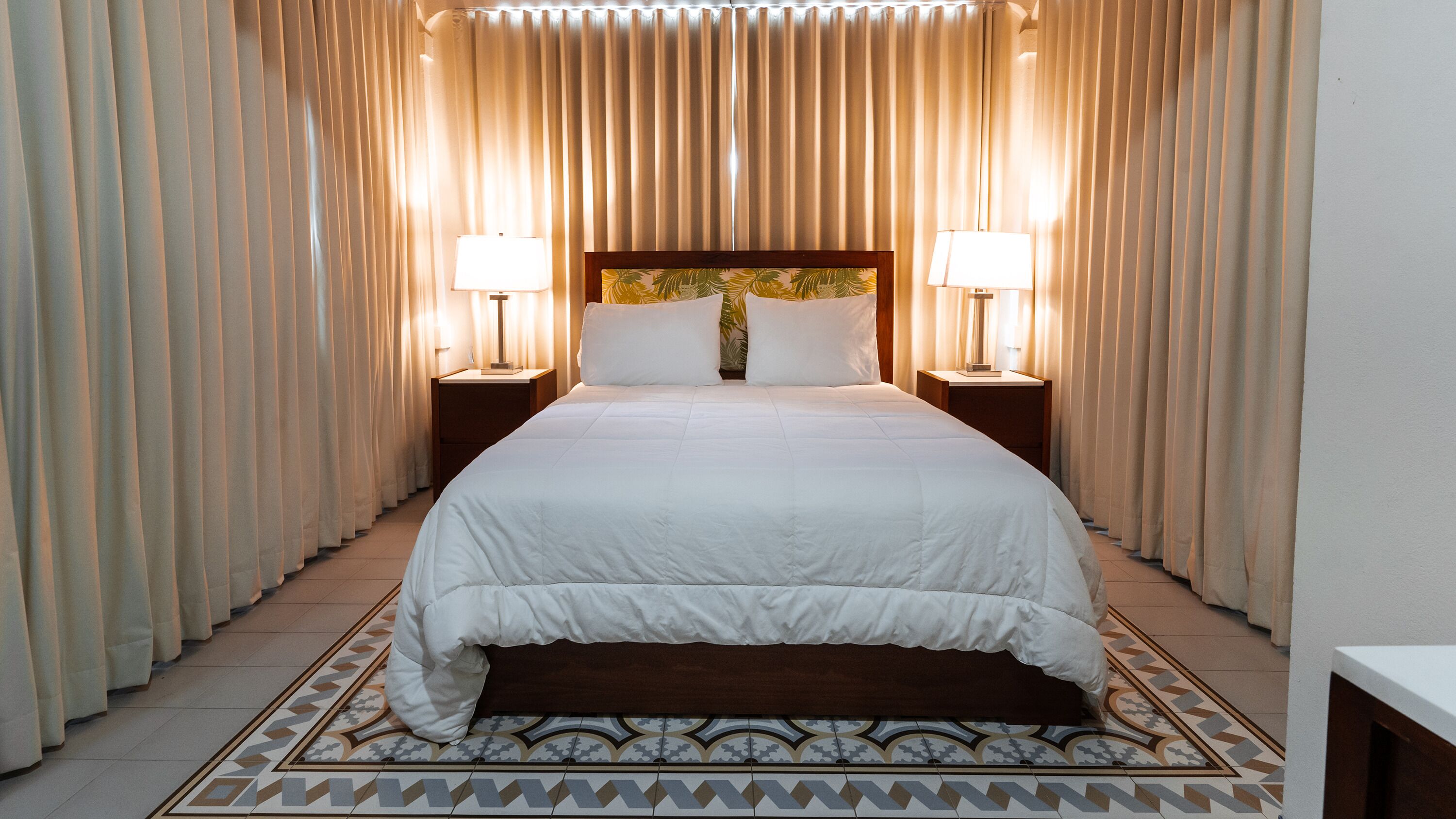 Uma cama grande com roupa de cama branca, ladeada por duas mesas de cabeceira com abajures, em um quarto com piso de azulejos estampados e cortinas translúcidas.