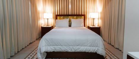 Uma cama grande com roupa de cama branca, ladeada por duas mesas de cabeceira com abajures, em um quarto com piso de azulejos estampados e cortinas translúcidas.