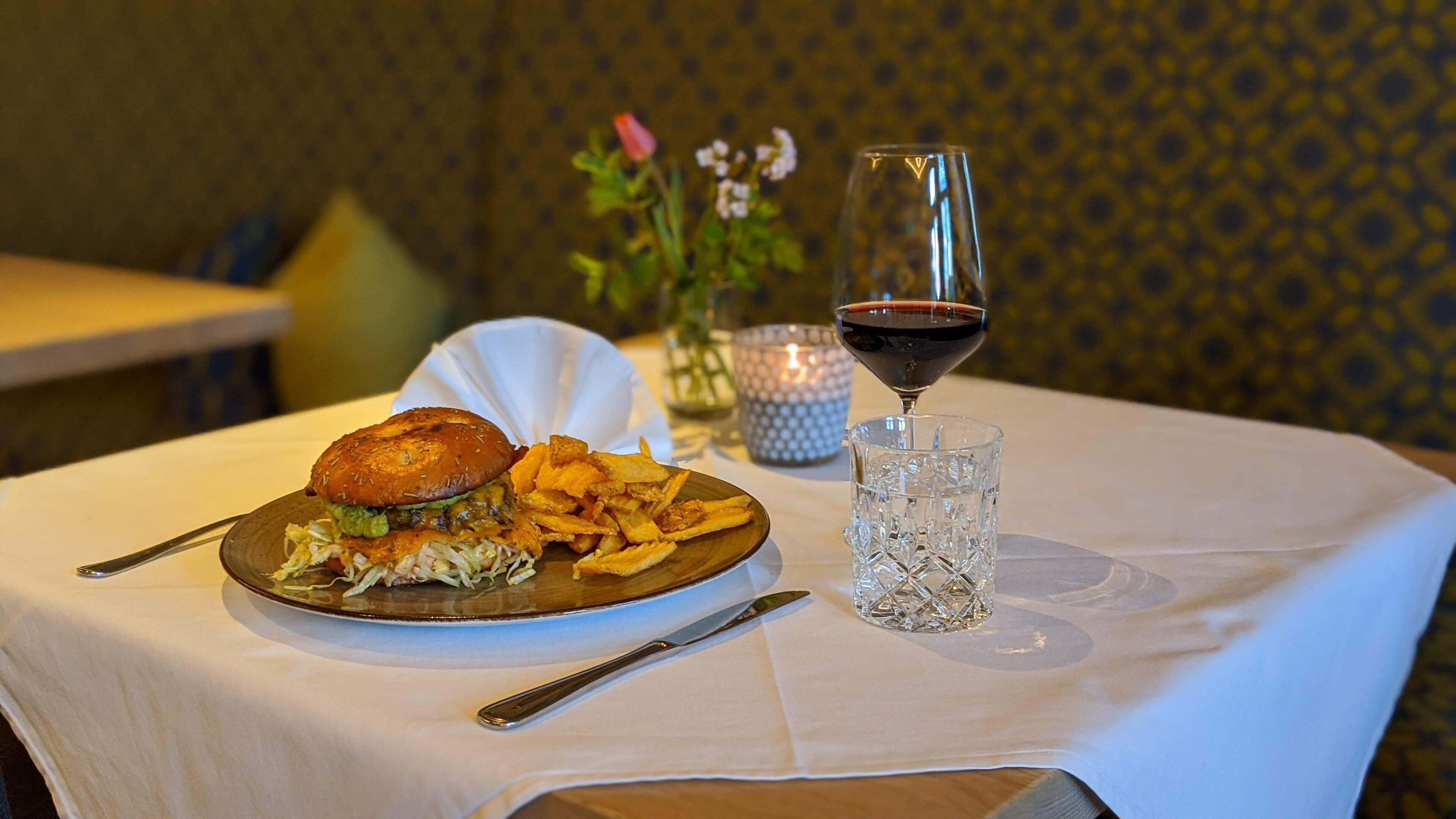 Uma mesa de jantar com um hambúrguer, batatas fritas, um copo de vinho tinto e uma vela.