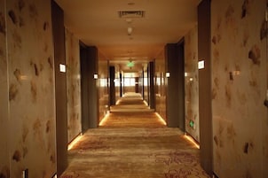 Corredor de hotel com piso em carpete, portas em ambos os lados e um longo trecho que leva a uma área iluminada no final.