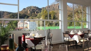 Restaurante com mesas preparadas para refeições, amplas janelas com vista para uma paisagem montanhosa e diversos garrafas sobre a mesa.