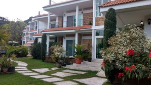 Um edifício residencial com vários andares, varandas, um caminho de jardim pavimentado e diversos vasos de plantas.