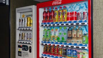 Máquina de vendas com diversas bebidas, incluindo Coca-Cola e Sapporo, e um refrigerador com a marca Coca-Cola, oferecendo uma variedade de bebidas.