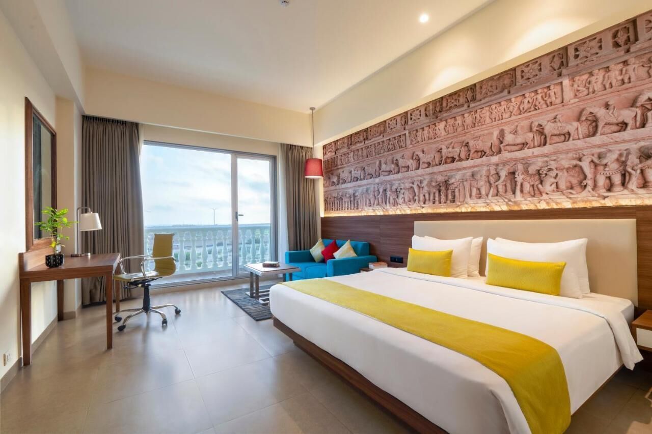 Quarto de hotel com cama grande, escrivaninha, poltrona, sofá e varanda com vista.