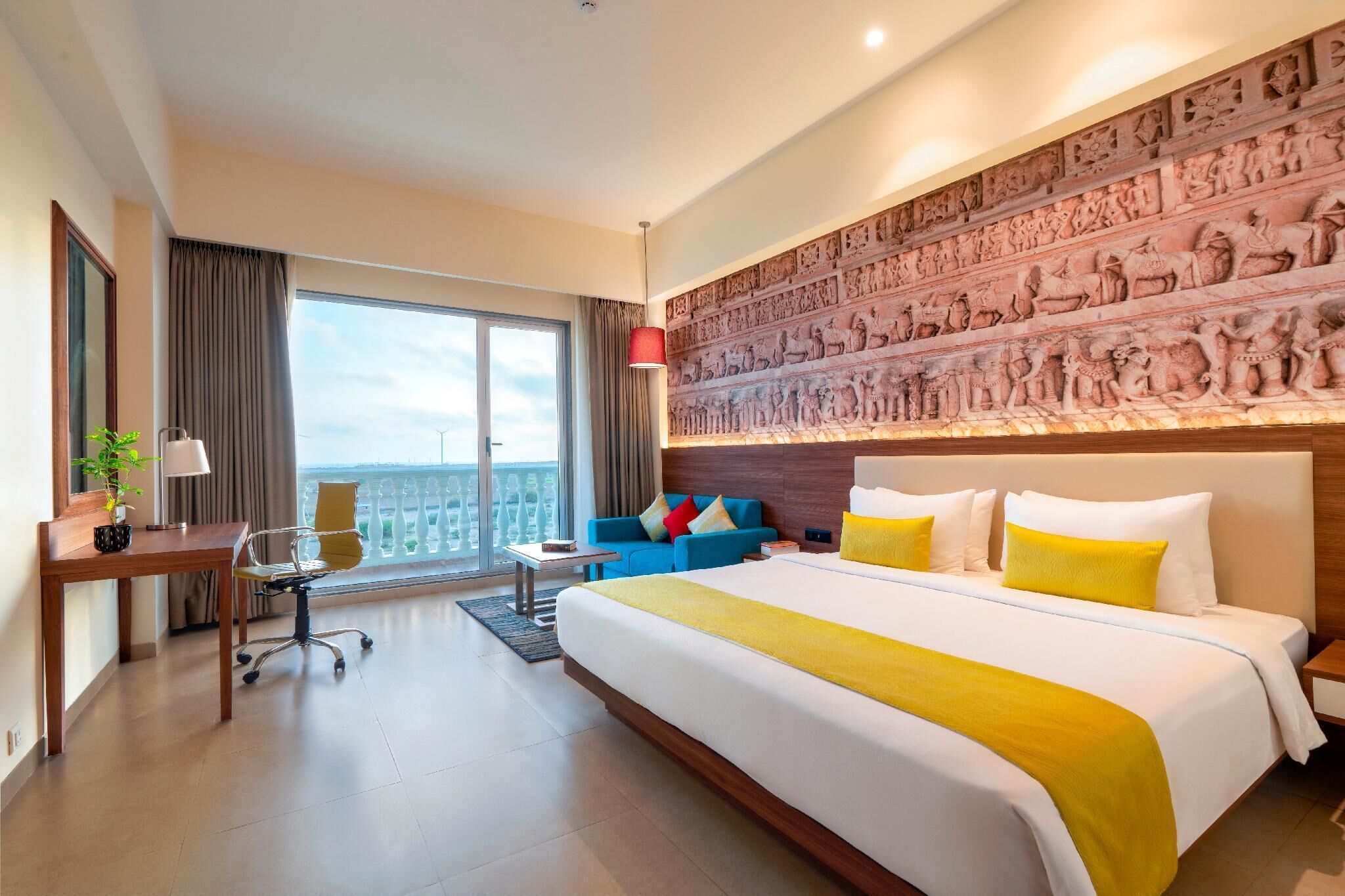 Quarto de hotel com uma cama grande, um sofá azul, uma escrivaninha com abajur e vista através de uma janela ampla.