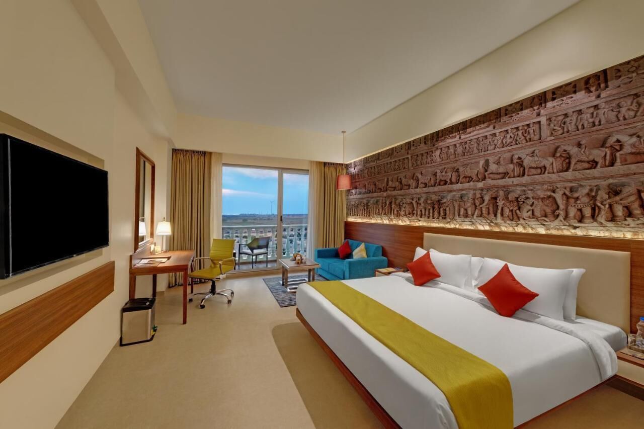 Quarto de hotel com uma cama grande, uma escrivaninha, uma cadeira, um sofá e uma parede com esculturas antigas.