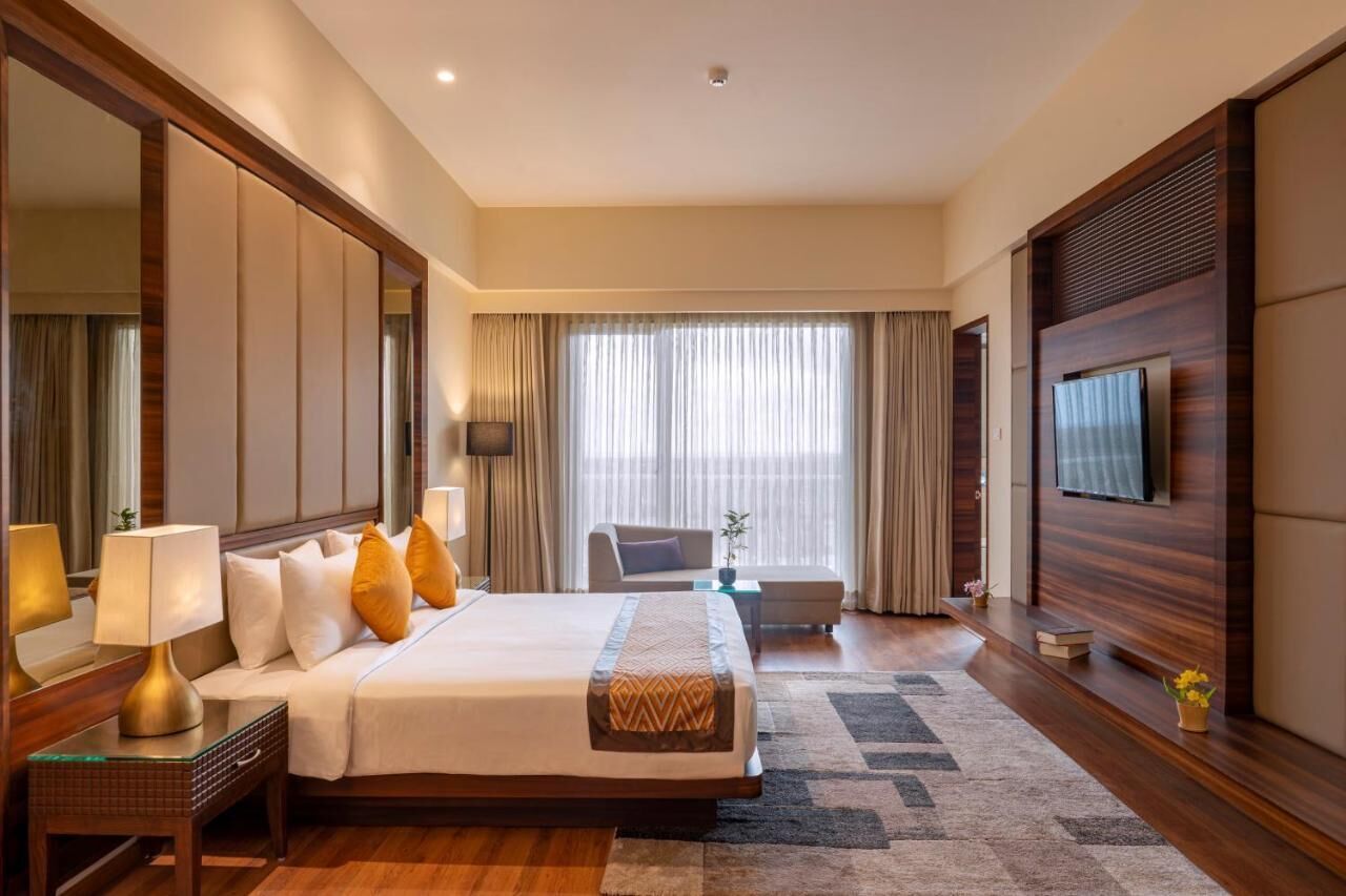 Quarto de hotel com uma cama grande, televisão na parede, escrivaninha com abajur e uma cadeira.