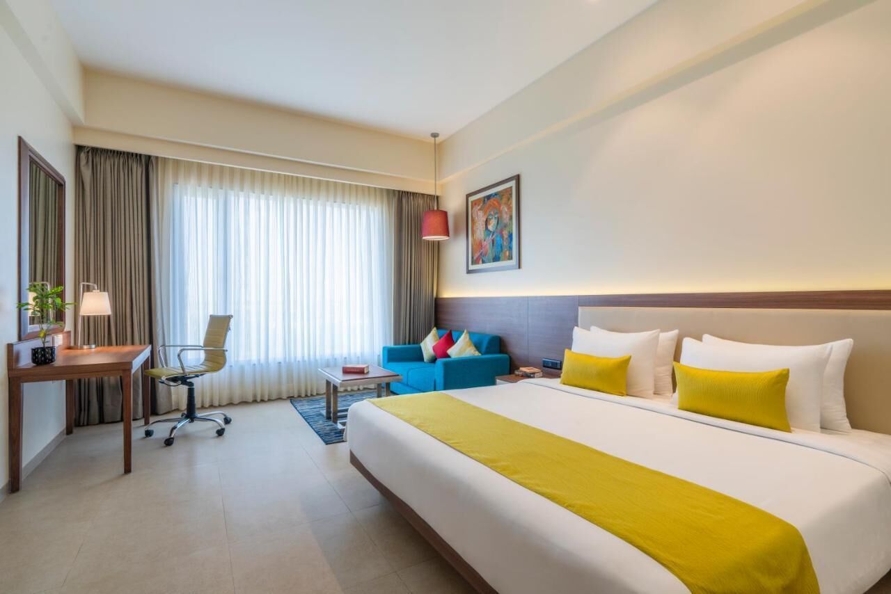 Quarto de hotel com uma cama grande, uma escrivaninha, uma cadeira, um sofá e uma janela com cortinas.