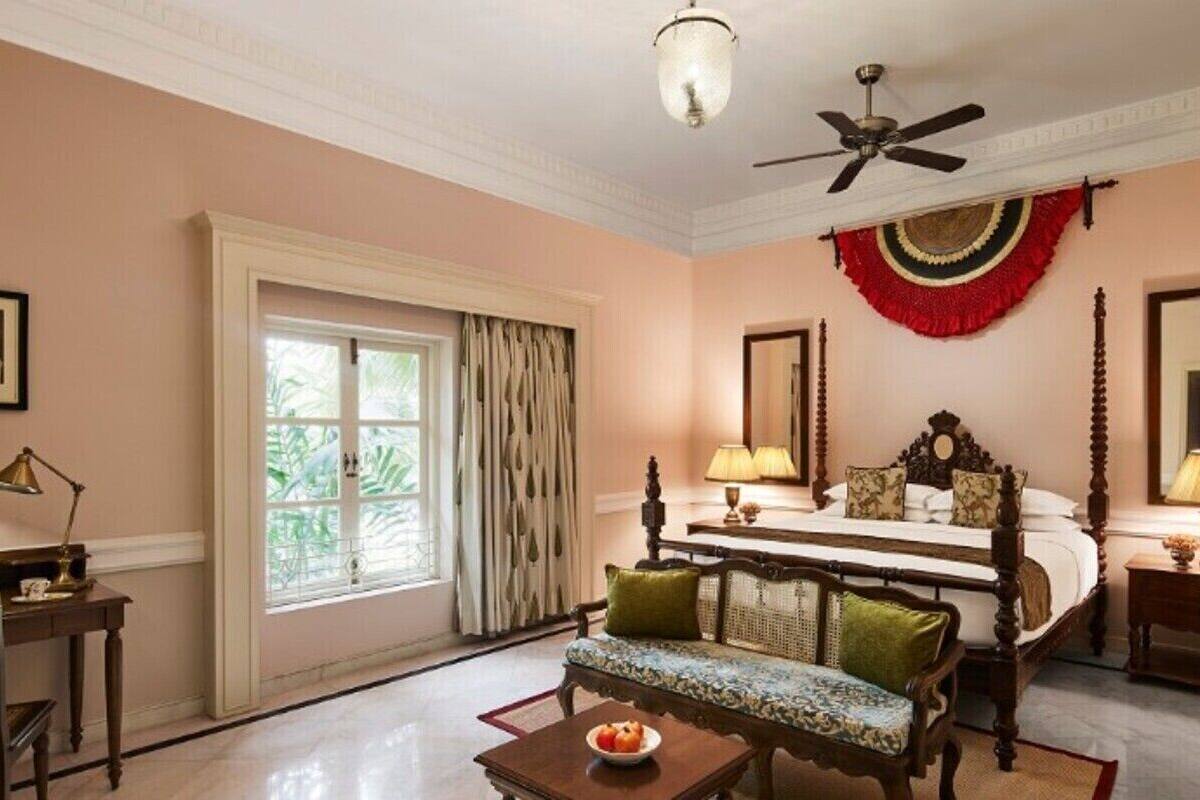 Quarto com cama, escrivaninha, cadeira, mesa com tigela, ventoinha no teto e janela com cortinas.