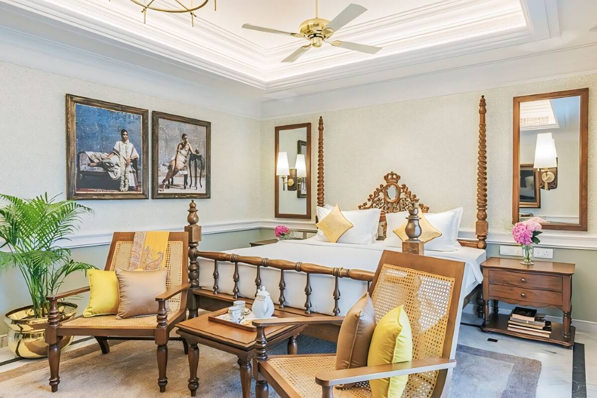 Quarto de hotel com uma cama grande, duas cadeiras, uma mesa, um espelho e quadros nas paredes.
