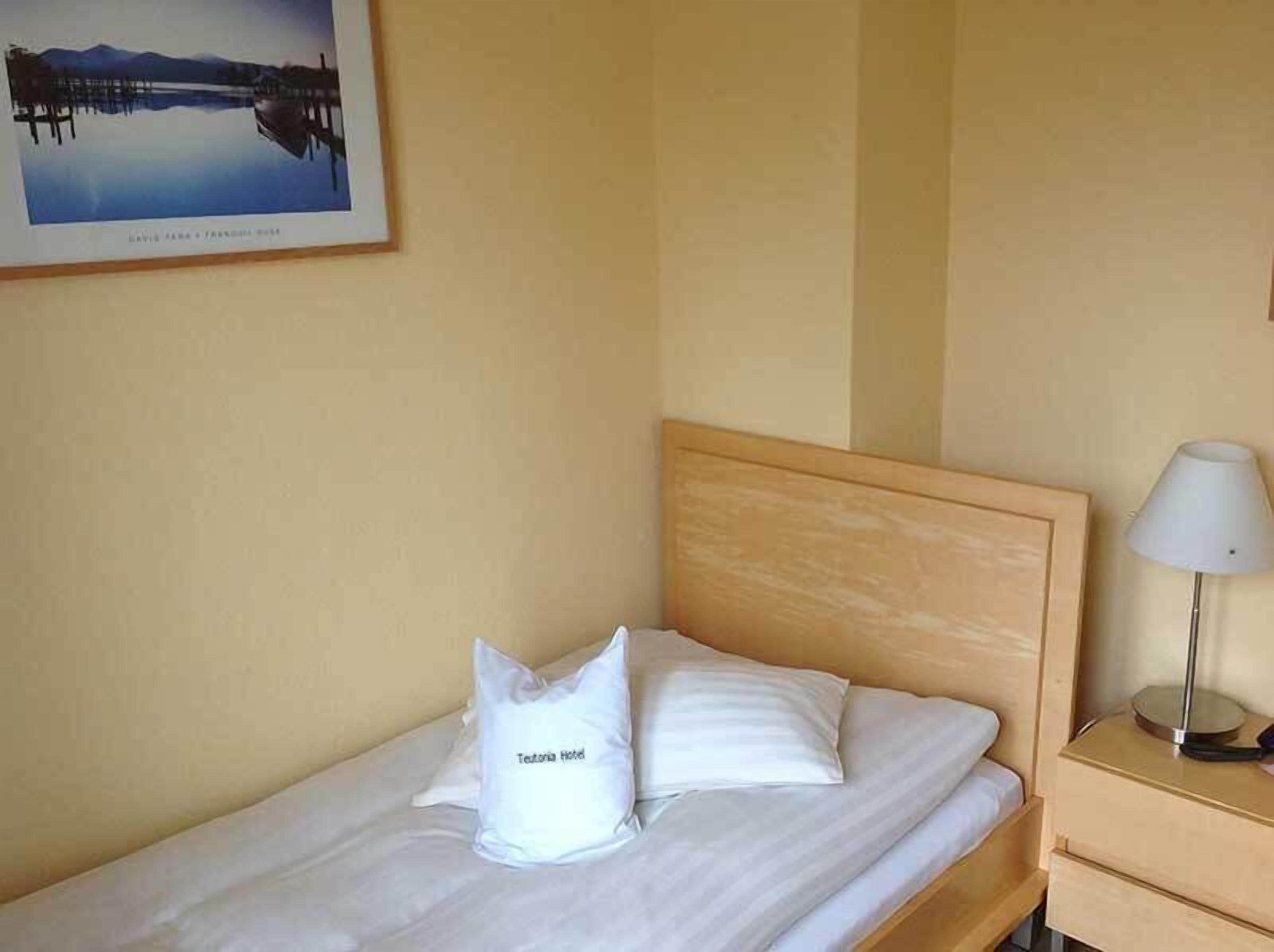 Uma cama de solteiro com cabeceira de madeira, duas almofadas, uma mesa de cabeceira com abajur e um quadro na parede.