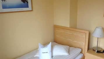 Uma cama de solteiro com cabeceira de madeira, duas almofadas, uma mesa de cabeceira com abajur e um quadro na parede.