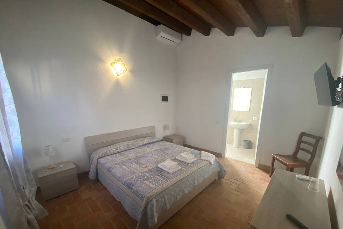 Quarto com cama, mesa de cabeceira e uma pequena escrivaninha com cadeira. Há uma janela no lado esquerdo e uma porta que dá para um banheiro.
