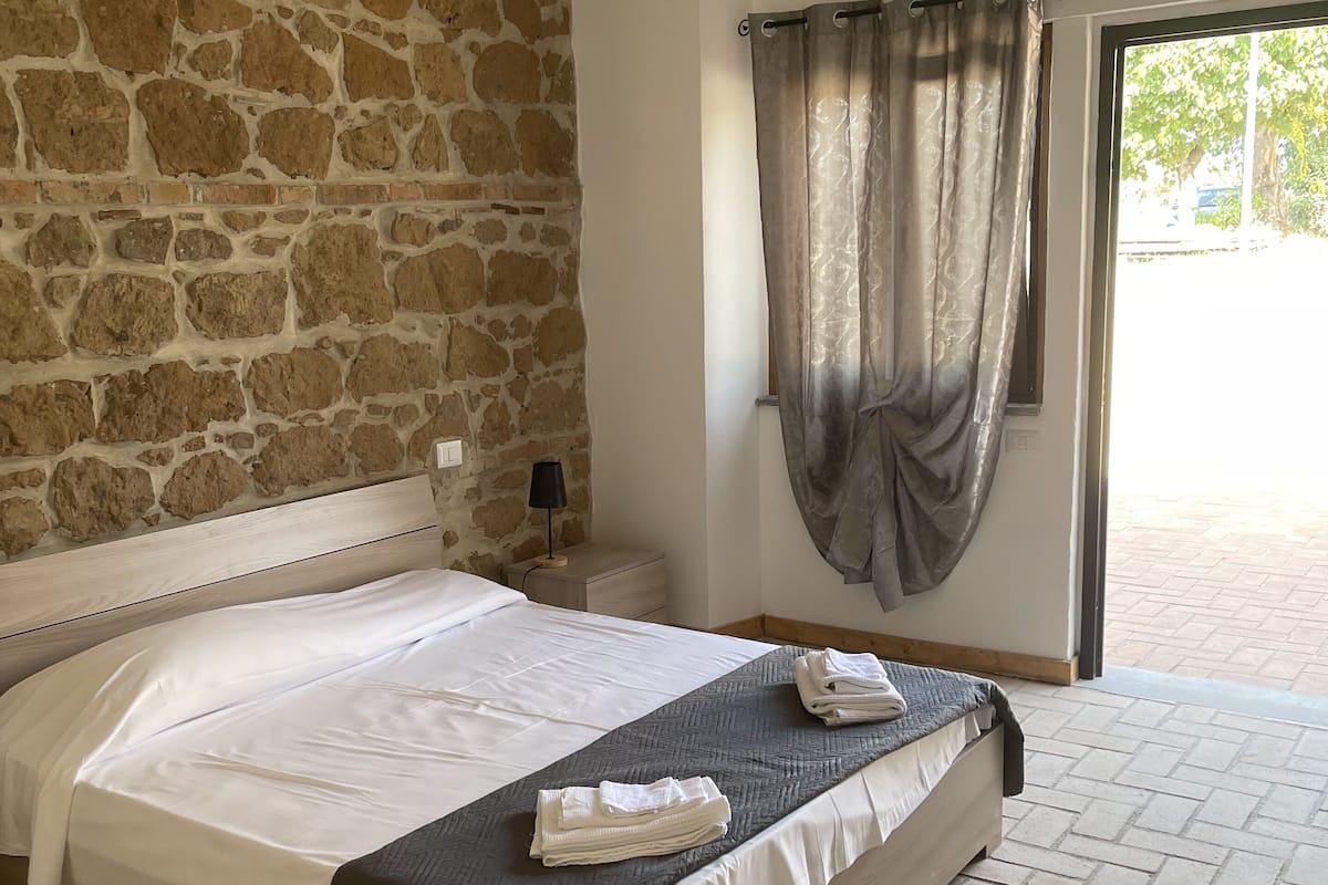 Quarto com parede de pedra, cama com roupa de cama branca e um espalhado cinzento, e janela com cortinas.